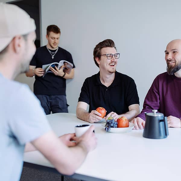 Vier Teammitglieder von mindtwo im Gespräch am Tisch, mit Kaffee und Obst, im Hintergrund liest jemand eine Zeitschrift