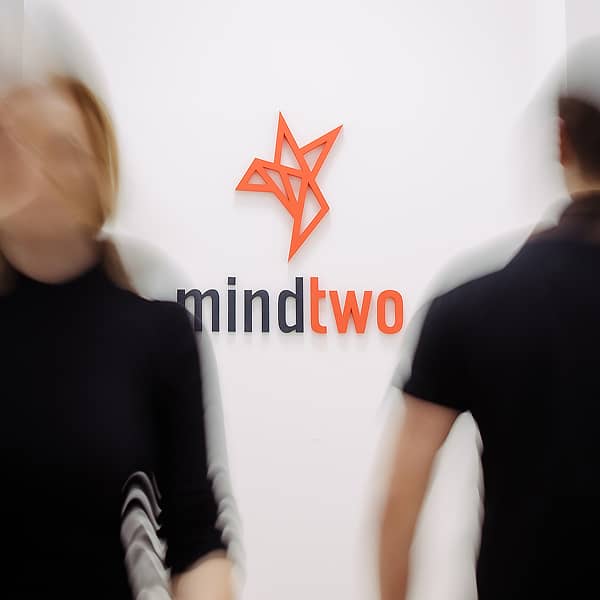 Unscharfe Aufnahme zweier vorbeigehender Personen vor dem mindtwo Logo an der Wand